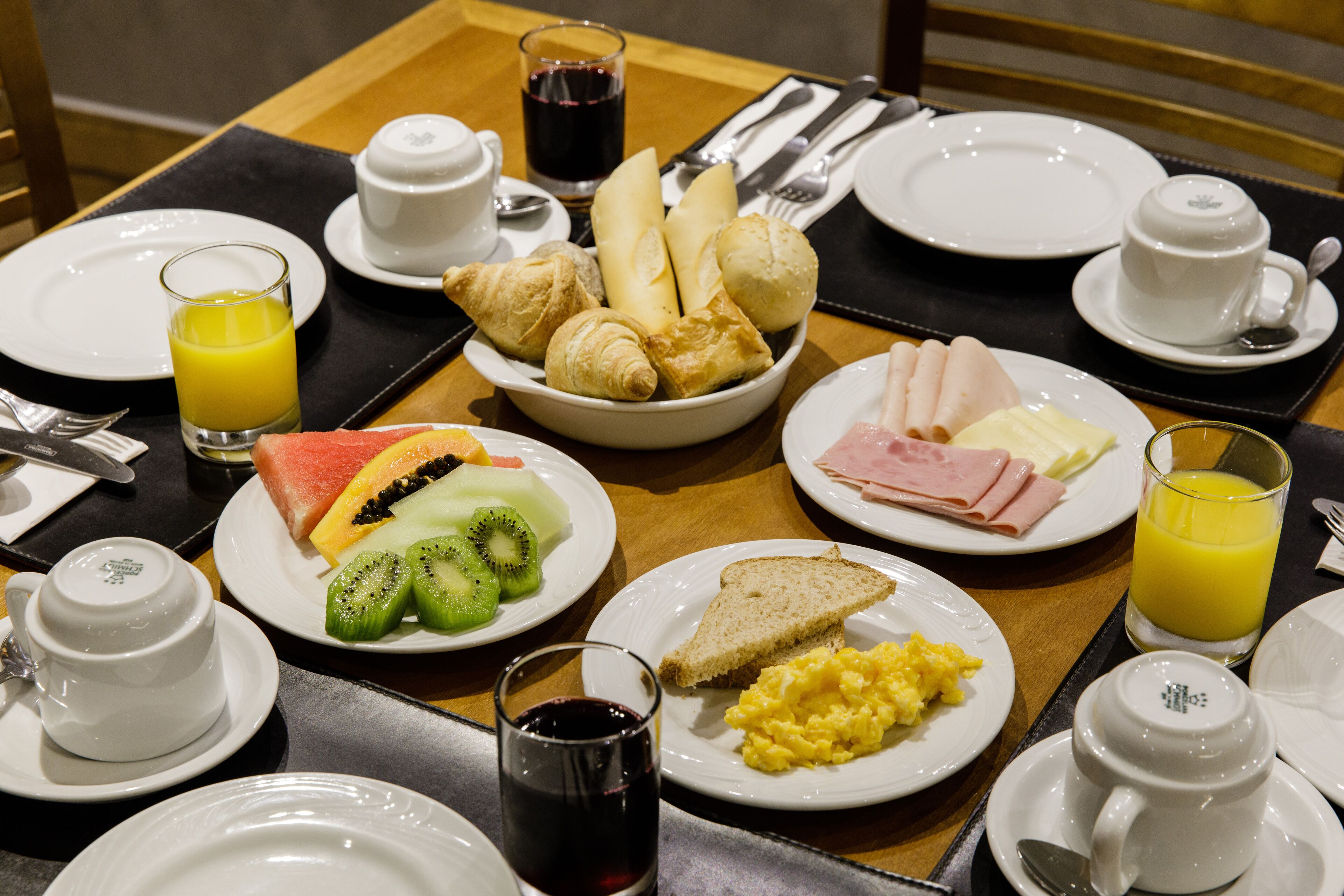 Mesa de café da manhã com pratos de comida, incluindo croissants, presunto, ovos e frutas, além de copos de suco e café.