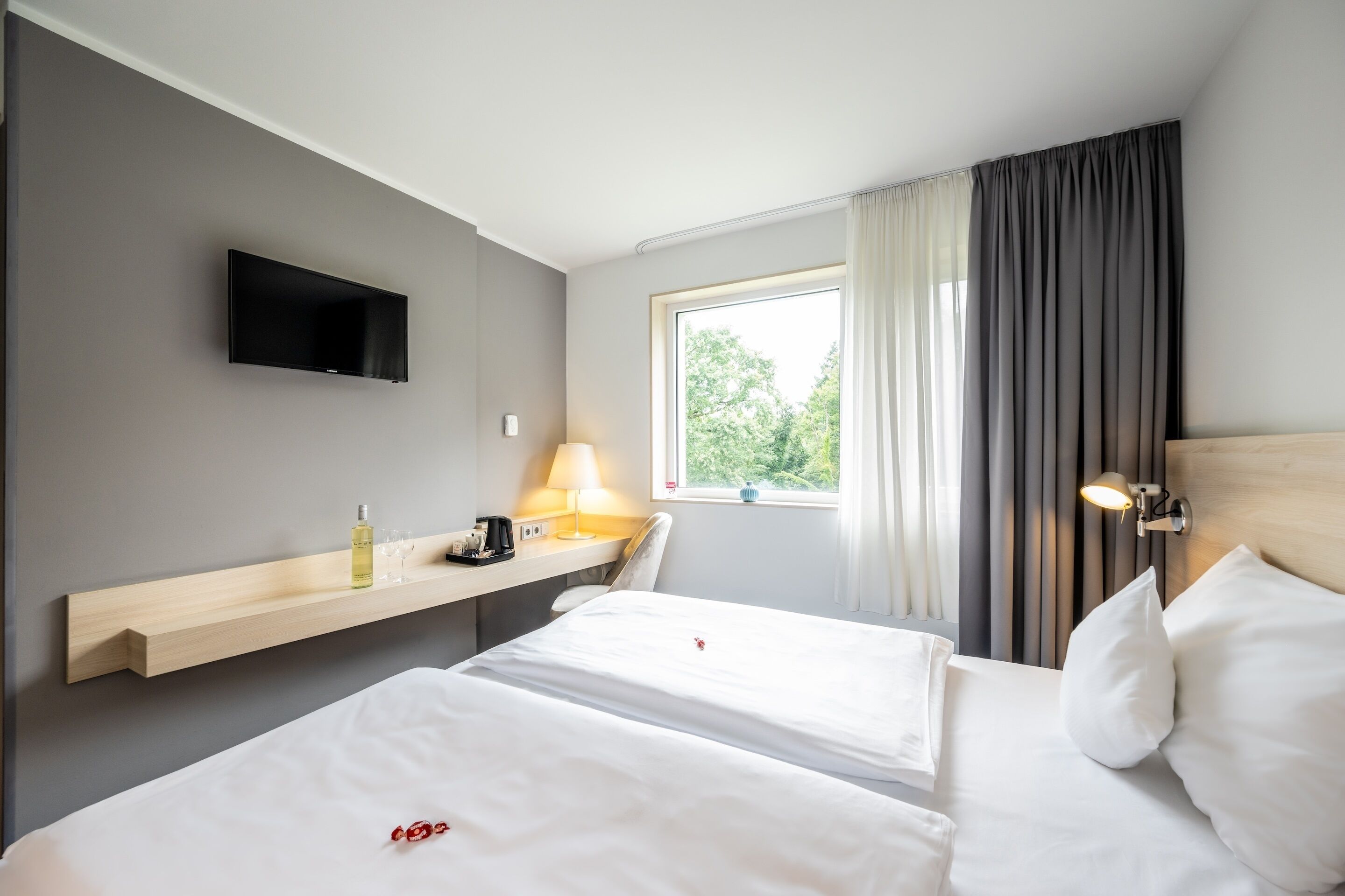 Quarto de hotel com duas camas, televisão presa na parede, mesa de cabeceira com abajur e janela com cortinas.