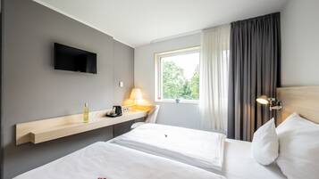 Quarto de hotel com duas camas, televisão presa na parede, mesa de cabeceira com abajur e janela com cortinas.