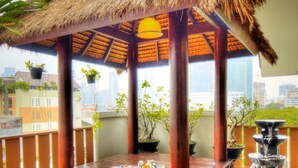 Um gazebo com telhado de palha, com mesa e cadeiras, plantas em vasos e um recurso de água em uma varanda com vista para a cidade.