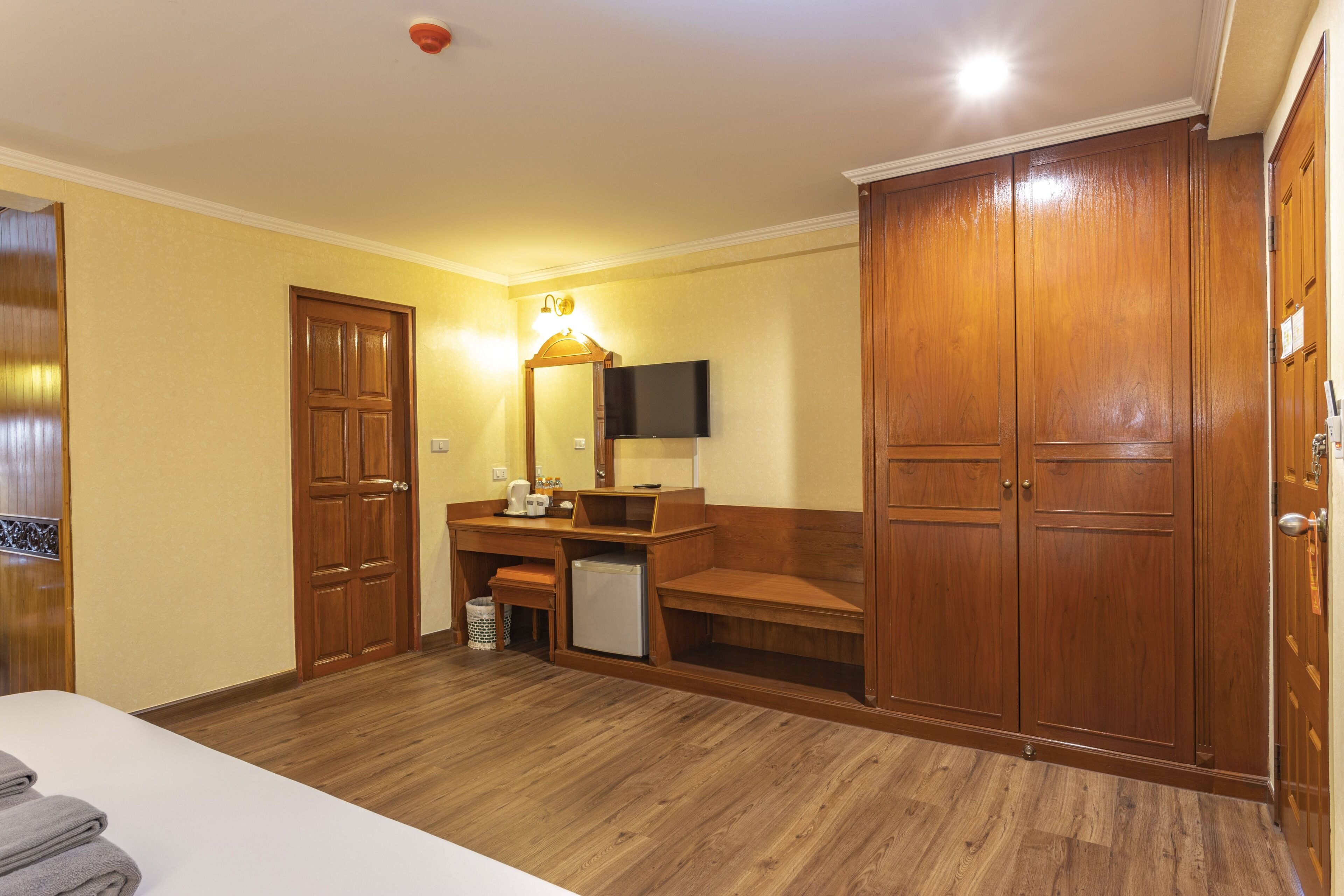 Quarto de hotel com piso de madeira, uma cama, escrivaninha com espelho, televisão e guarda-roupa.