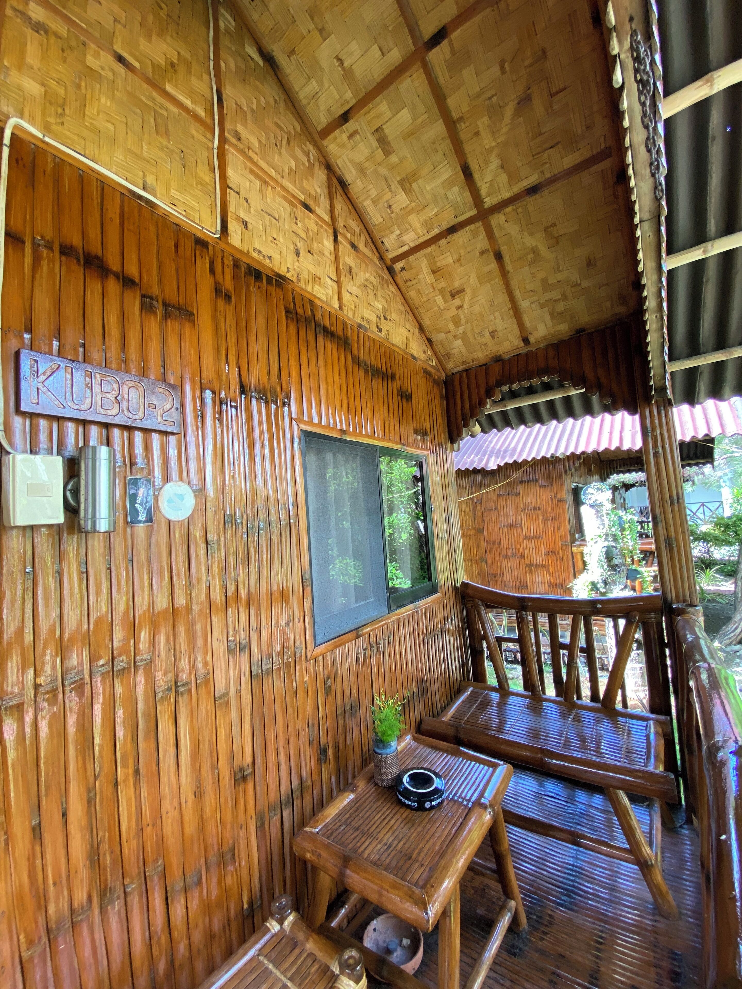 Uma cabana de madeira com uma placa indicando "KUBO-2", uma janela, uma mesa pequena com uma tigela e um banco.