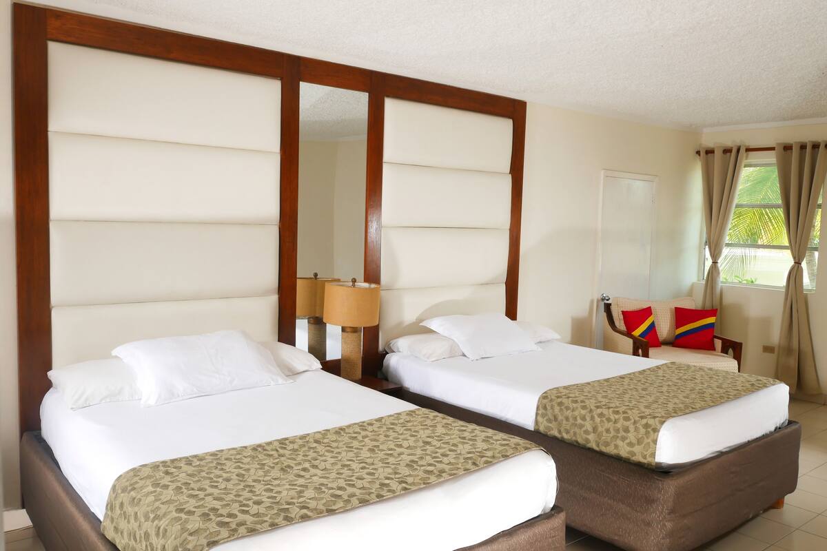 Quarto de hotel com duas camas, um espelho grande, mesa de cabeceira com abajur e janela com cortinas.