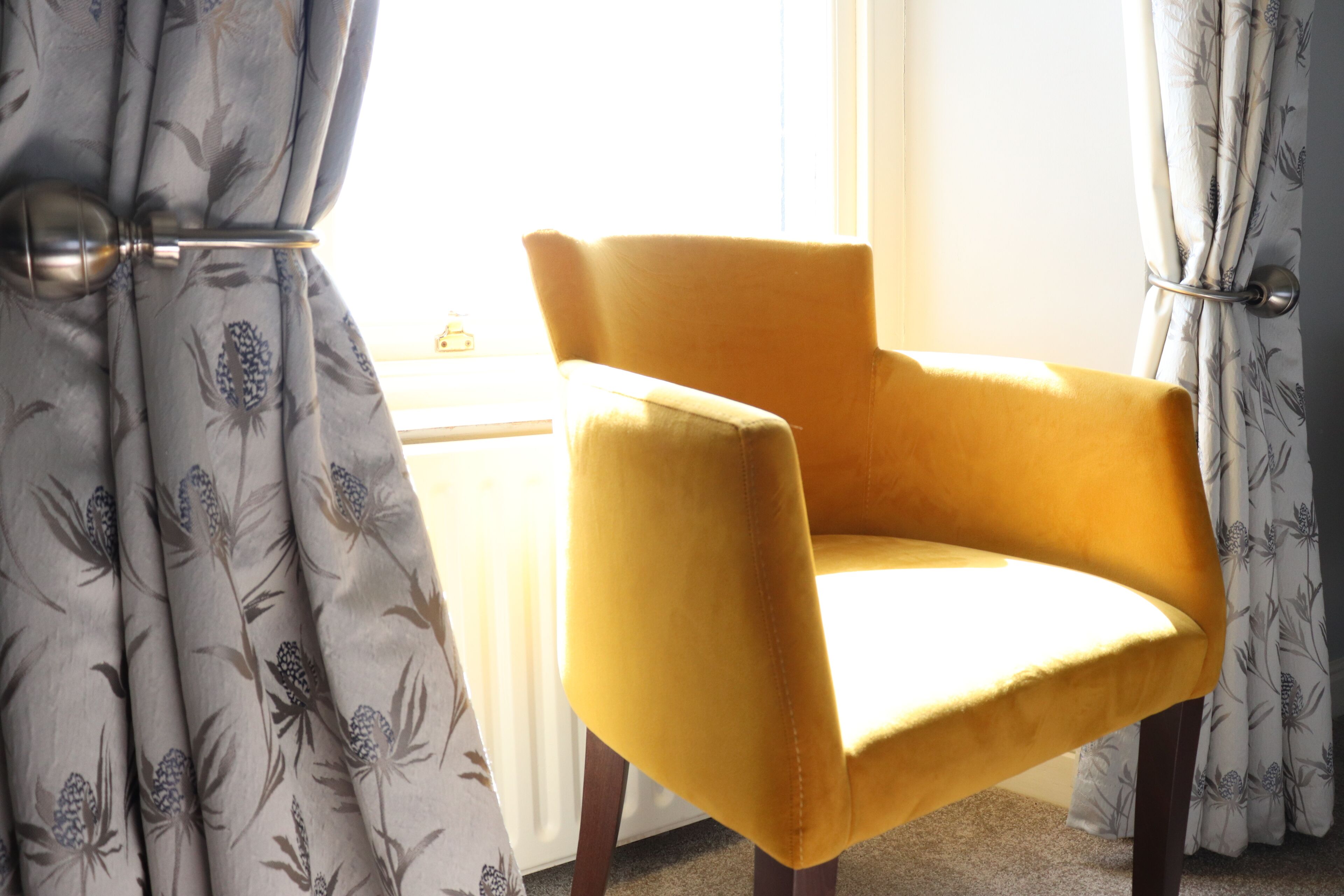 Quarto com poltrona amarela perto de uma janela, cortinas florais e radiador.