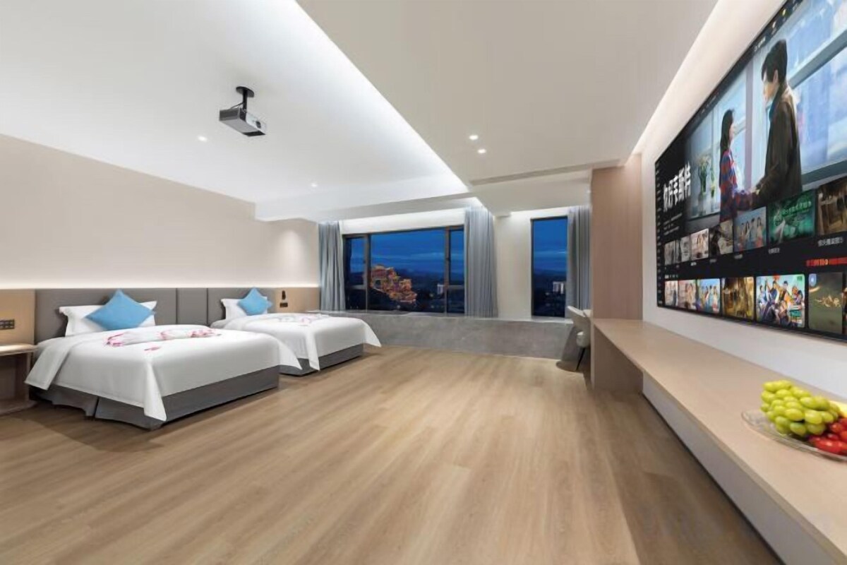 Quarto de hotel moderno com duas camas, uma janela grande e uma TV fixada na parede exibindo diversos conteúdos.