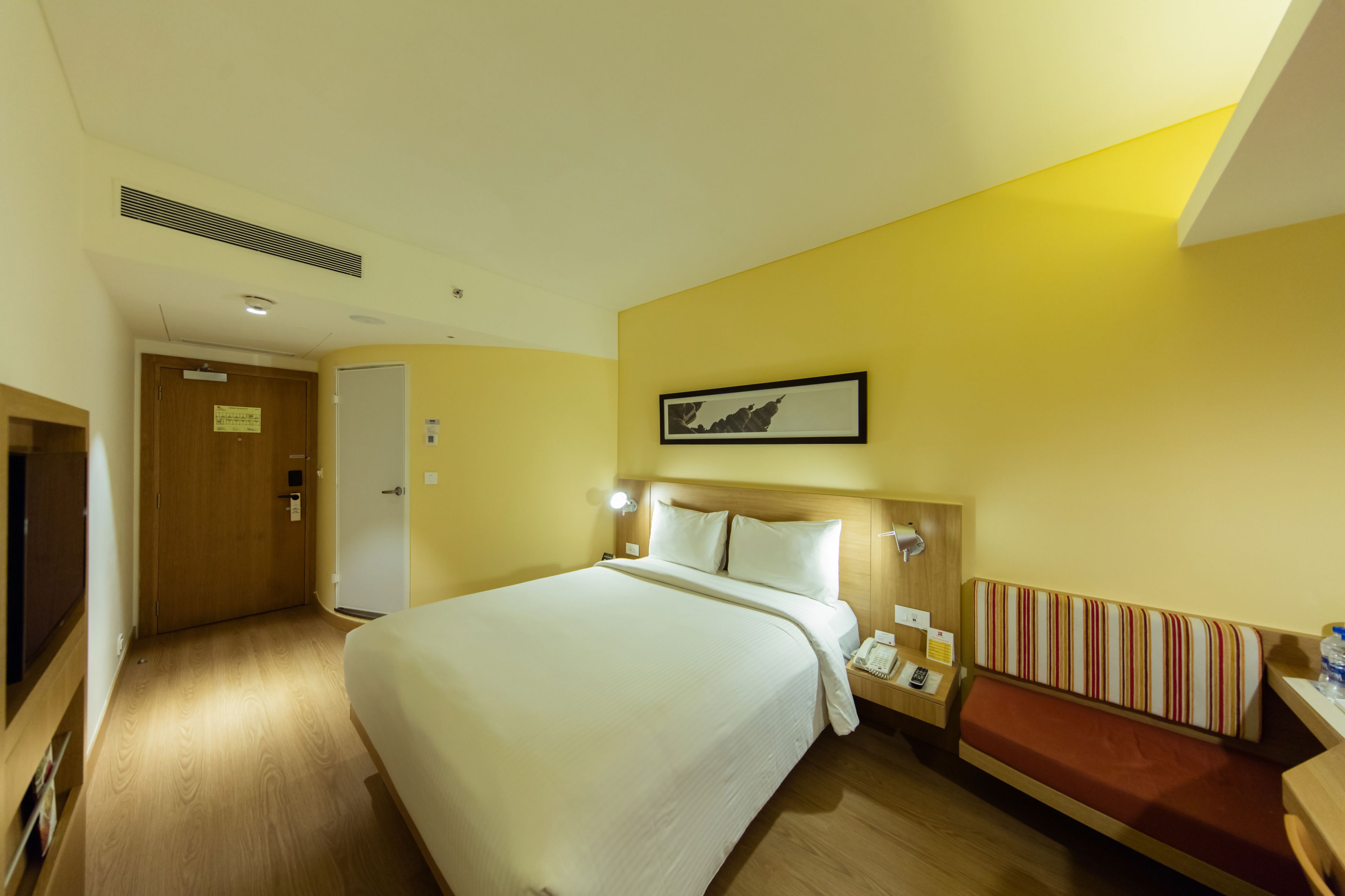 Quarto de hotel com uma cama grande, uma mesinha de cabeceira com abajur, uma pequena mesinha com telefone, um banquinho com almofada listrada e um quadro na parede.