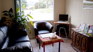 Uma sala de estar com um sofá de couro preto, um armário de madeira, uma mesinha com toalha xadrez, uma escrivaninha com monitor e uma janela grande com vista para o verde lá fora.