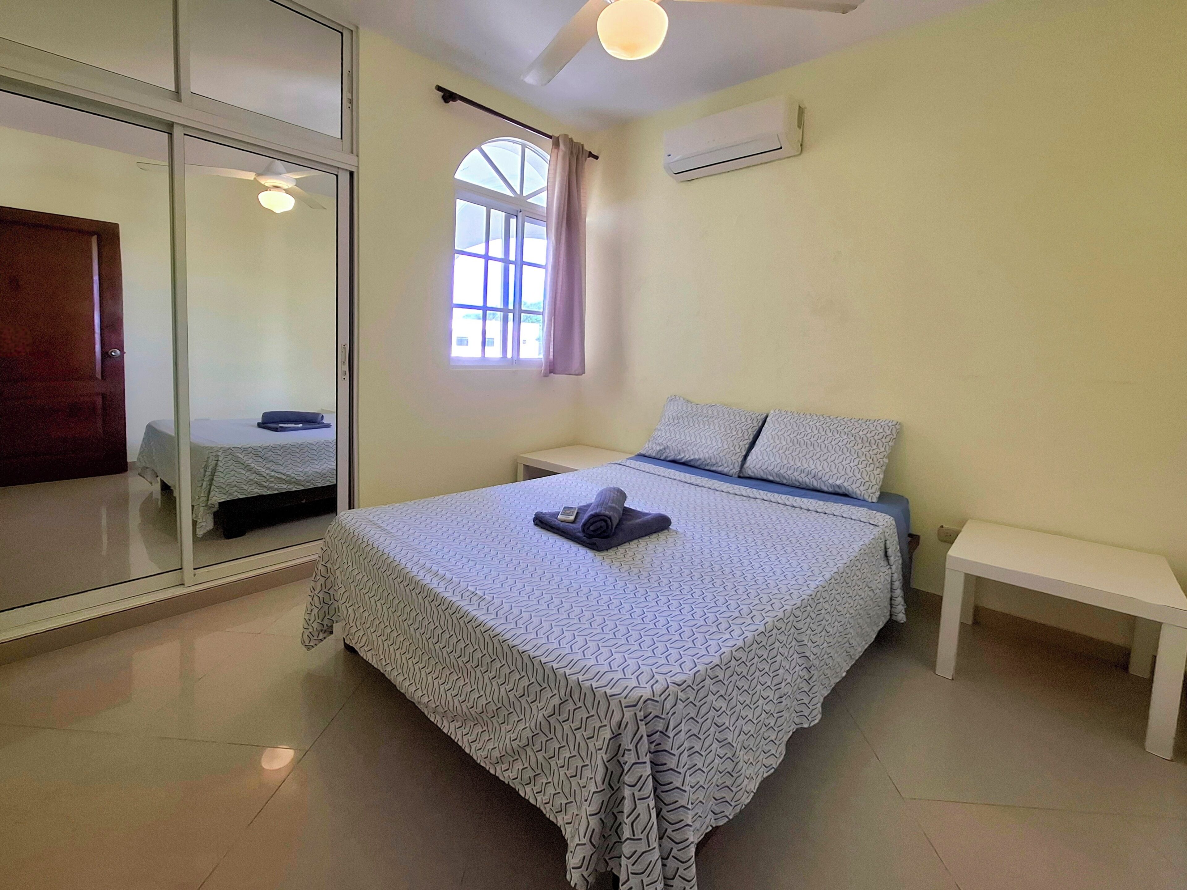 Quarto com cama, mesa de cabeceira, espelho, janela com cortinas e ar condicionado.