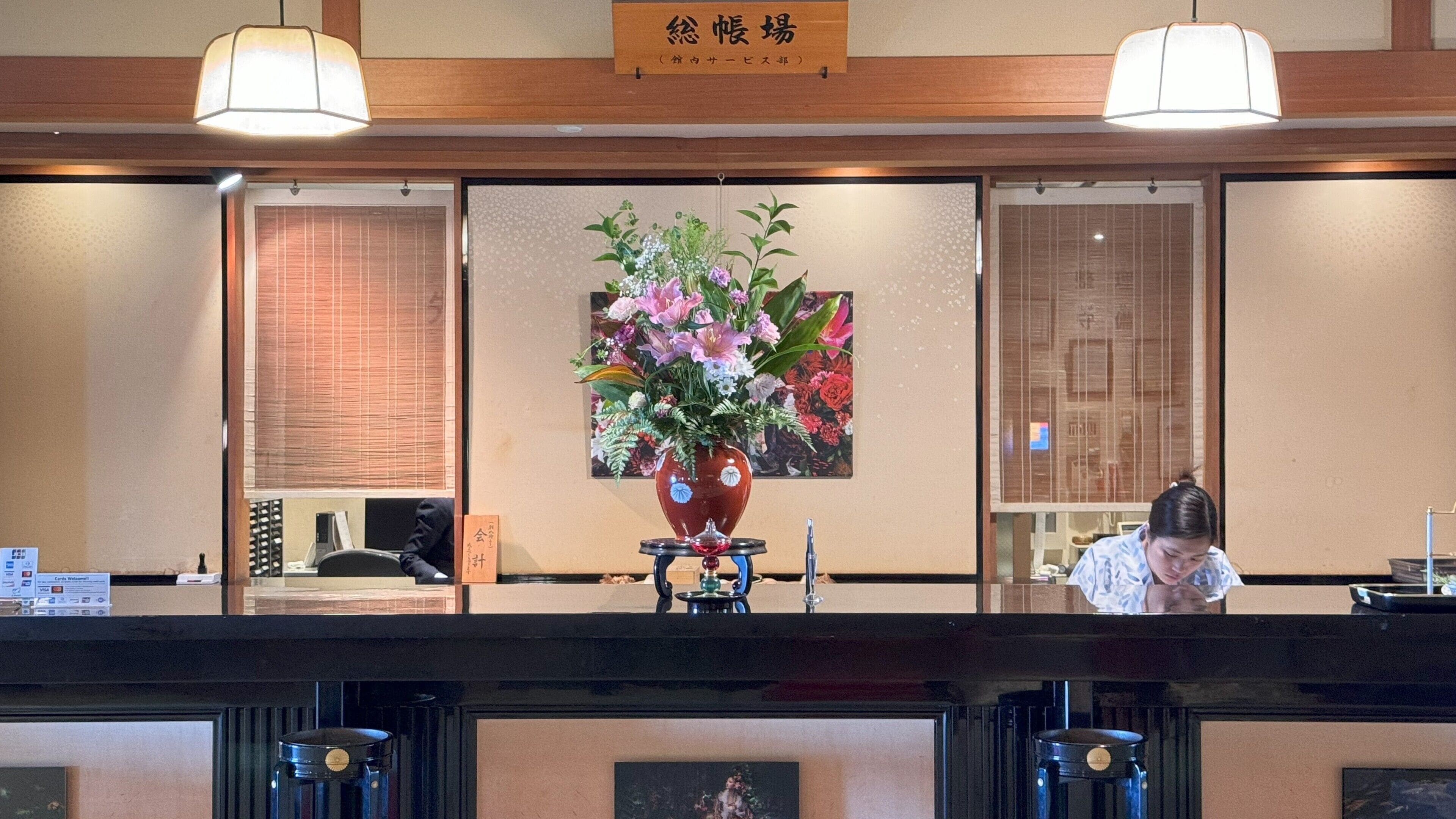 Uma receção com um grande vaso de flores, uma placa de madeira em cima e uma pessoa atrás do balcão.