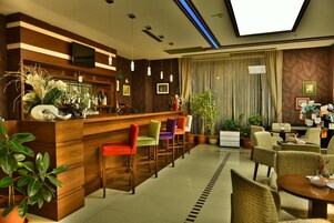 Um hall de hotel com balcão de recepção de madeira, banquetas coloridas, plantas em vasos e uma área de estar com cadeiras de vime.