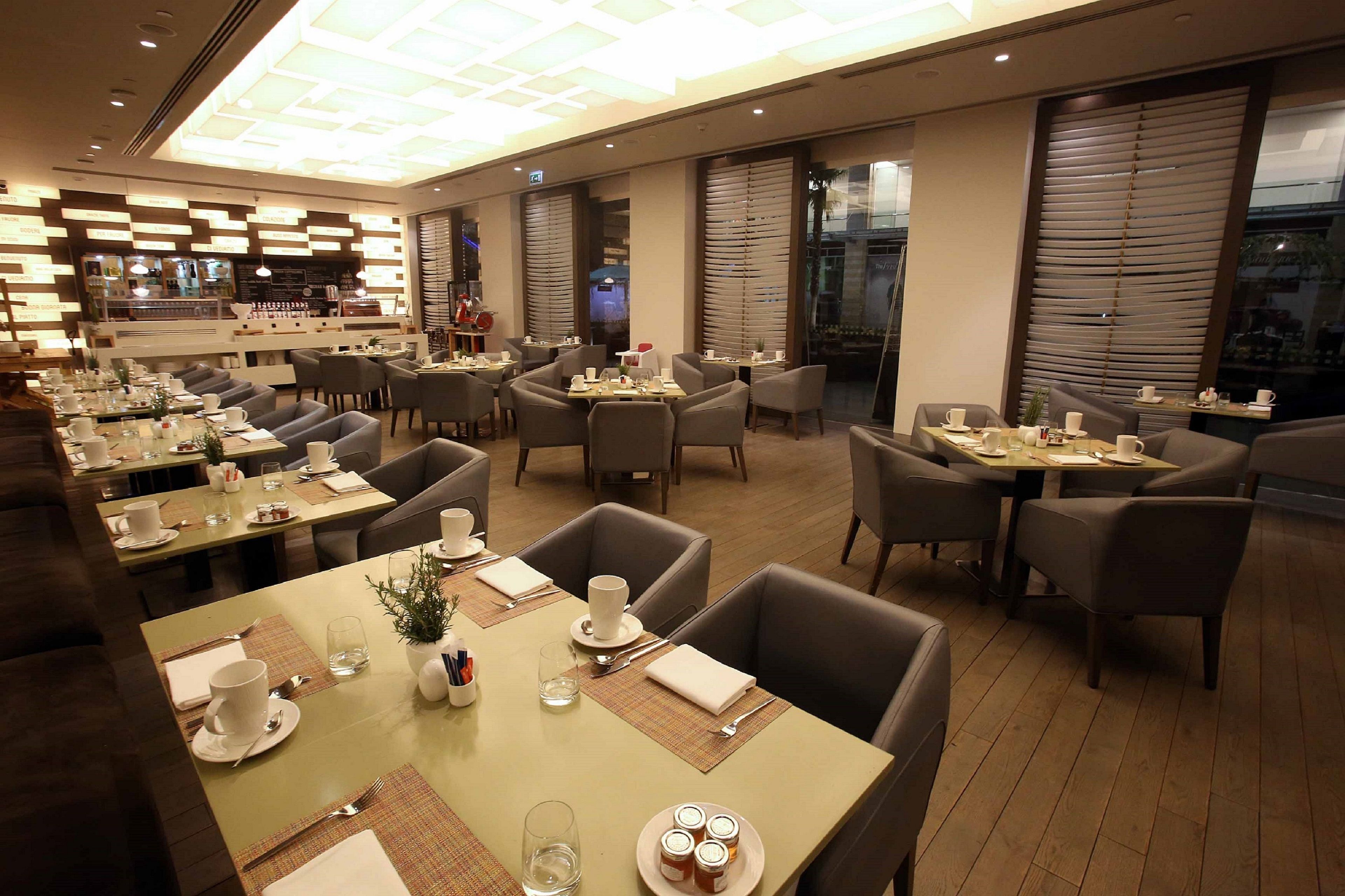 Um restaurante moderno com várias mesas e cadeiras, um bar e grandes janelas com persianas.