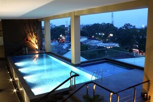 Piscina interior moderna com paredes de vidro transparente, proporcionando vista dos edifícios e árvores ao redor.