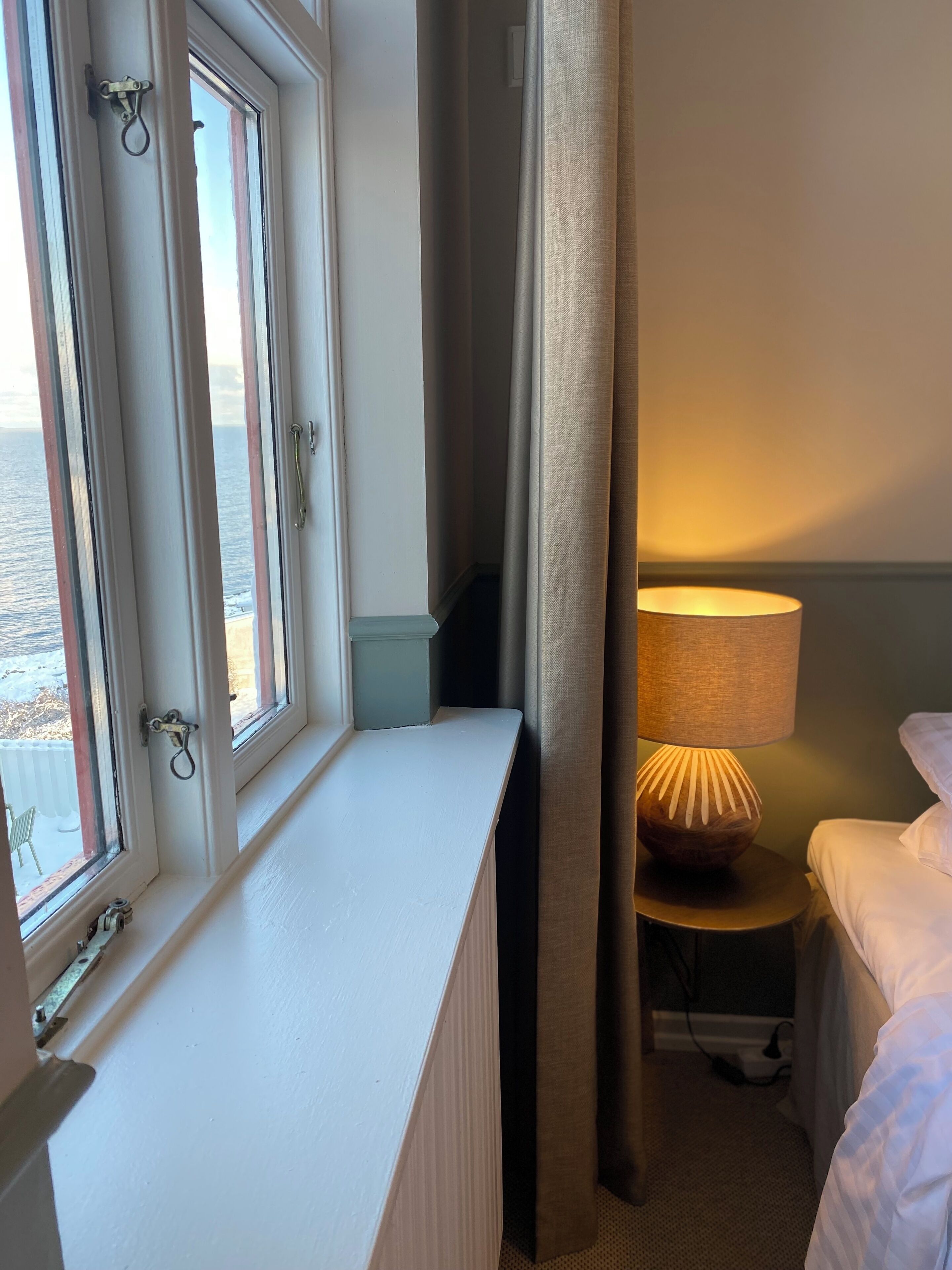 Quarto de hotel com janela, mesa de cabeceira e uma luminária, além de uma cama.