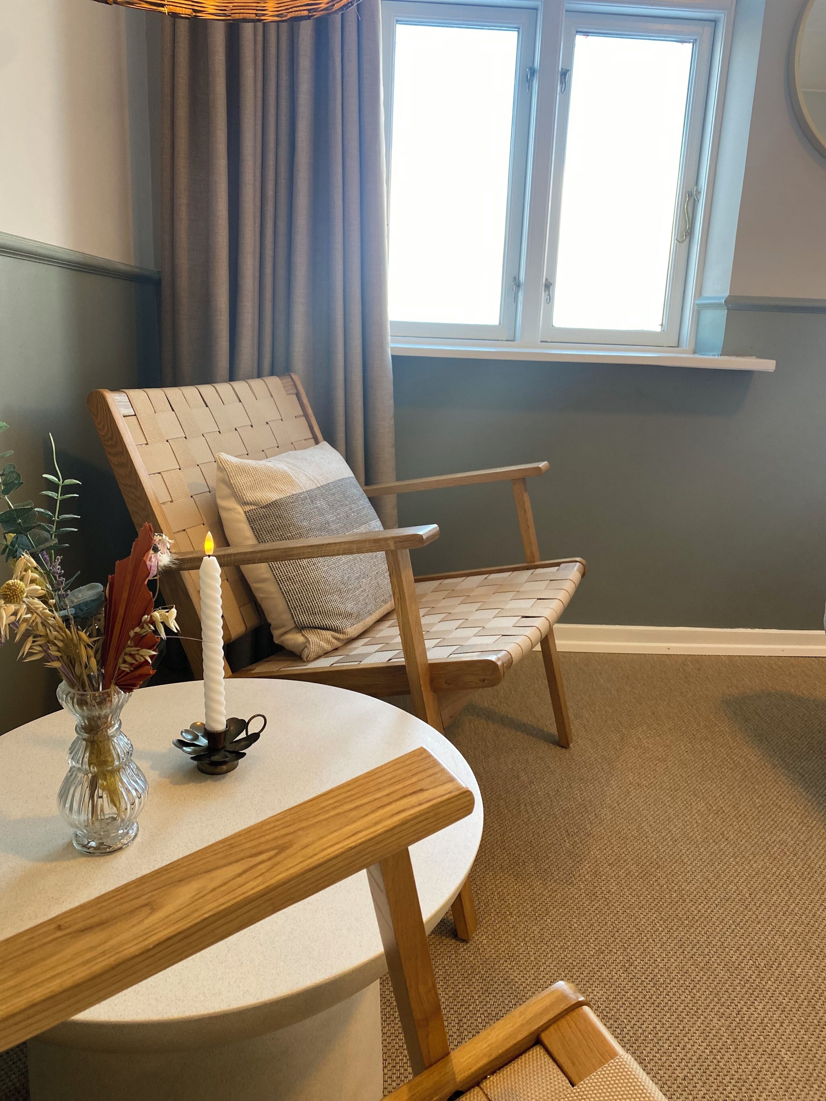 Um quarto acolhedor com uma cadeira de madeira, uma mesa redonda, um vaso com flores secas e uma vela.