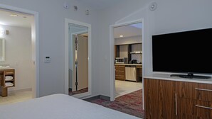 Quarto de hotel com cama, televisão, armário de madeira e cozinha visível através de uma porta aberta.