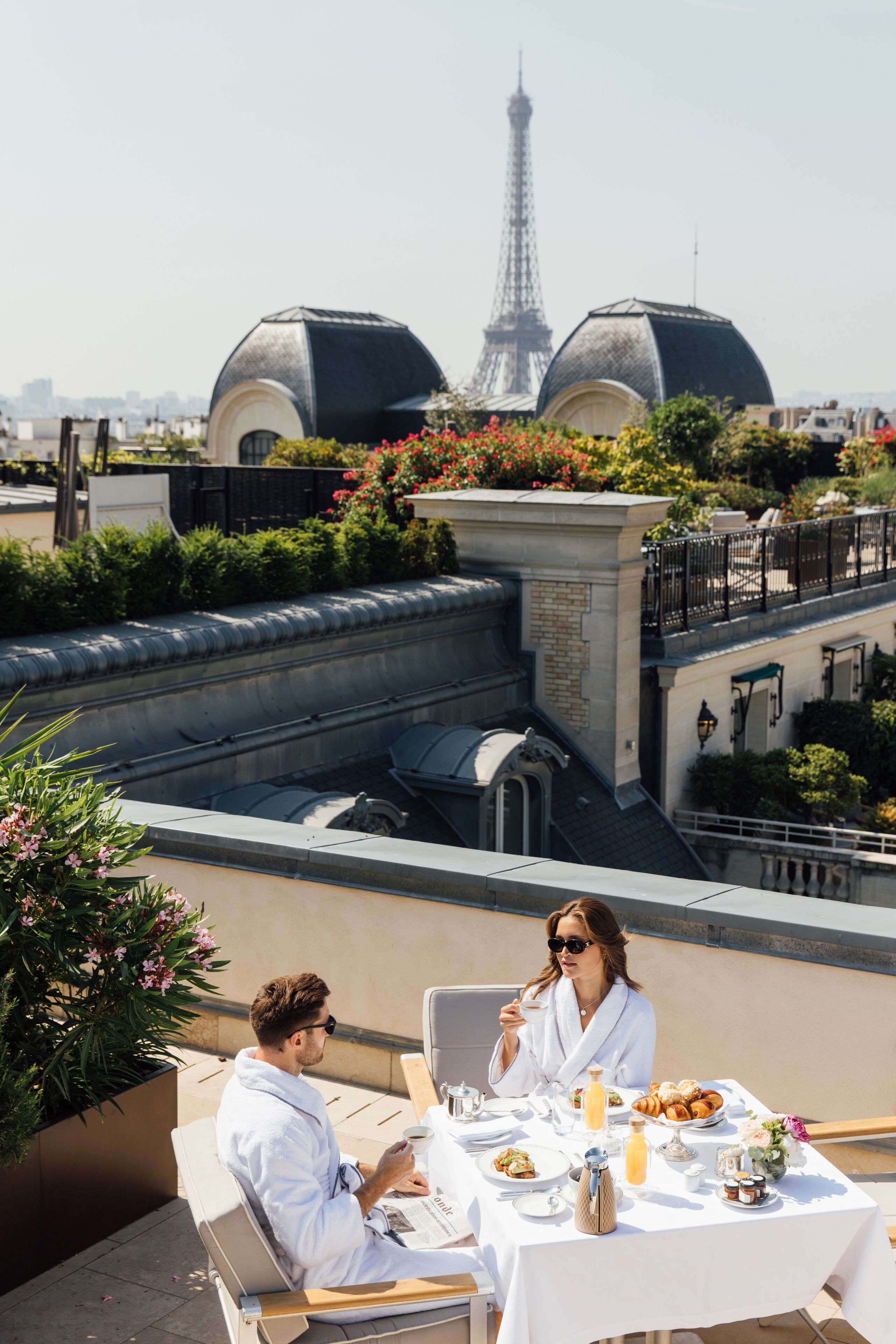 Uma área de refeições no terraço com vista para a Torre Eiffel, com uma mesa posta para o café da manhã e uma pessoa lendo um jornal.