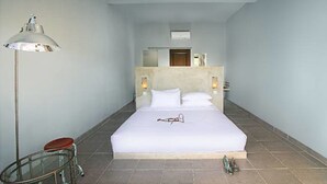Quarto de hotel com uma cama grande, uma mesinha de cabeceira, um abajur e um banquinho.
