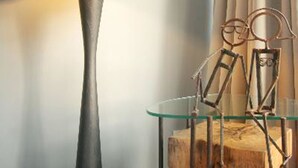 Uma luminária moderna com cúpula texturizada, uma escultura de madeira sobre uma mesa de vidro e uma cortina ao fundo.