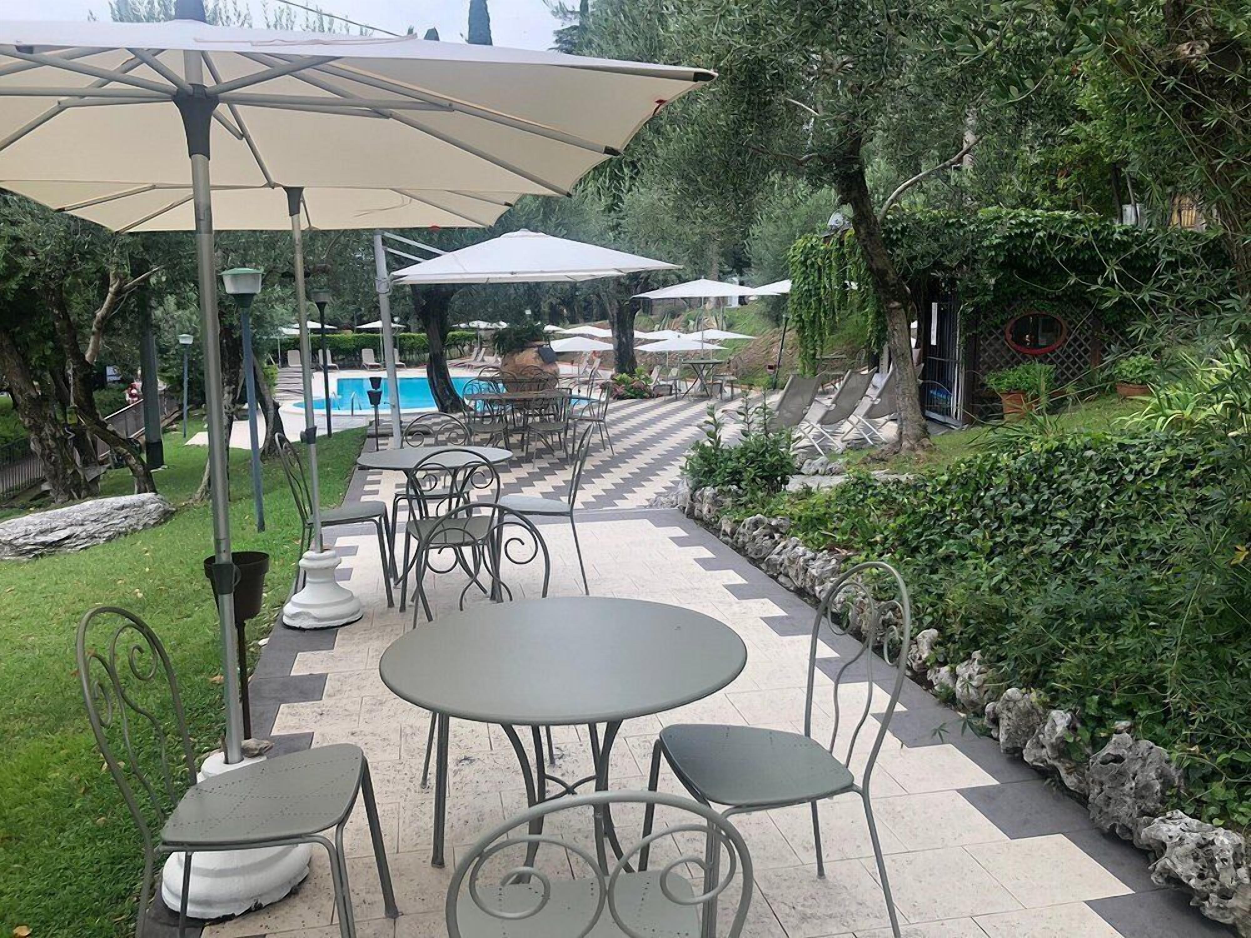 Uma área de pátio ao ar livre com mesas e cadeiras redondas, guarda-sóis e uma piscina ao fundo.