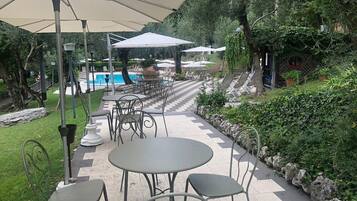 Uma área de pátio ao ar livre com mesas e cadeiras redondas, guarda-sóis e uma piscina ao fundo.