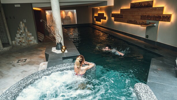 Piscina interior com área de hidromassagem, piso revestido em azulejo e parede de madeira com padrão em cruz.