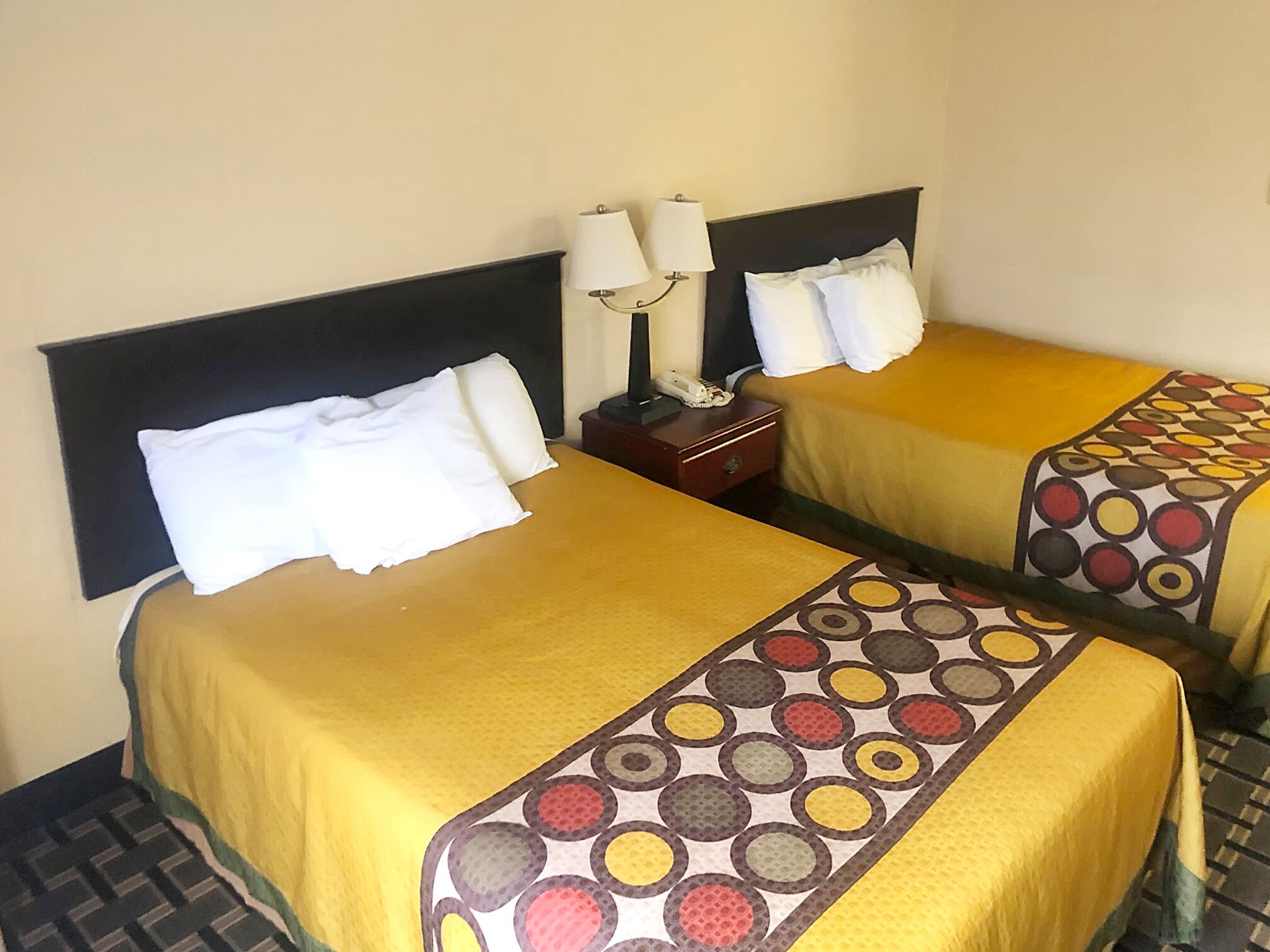Quarto de hotel com duas camas, cada uma com cabeceira, mesas de cabeceira, luminárias e cobreleito estampado.