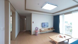 Quarto de hotel moderno com uma janela grande, uma televisão de tela plana na parede, uma mesa de centro de madeira e uma pequena estante com objetos decorativos.
