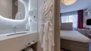 Quarto de hotel moderno com um espelho grande, pia embutida, roupão pendurado em um porta-toalhas, cama com cabeceira e televisão na parede.