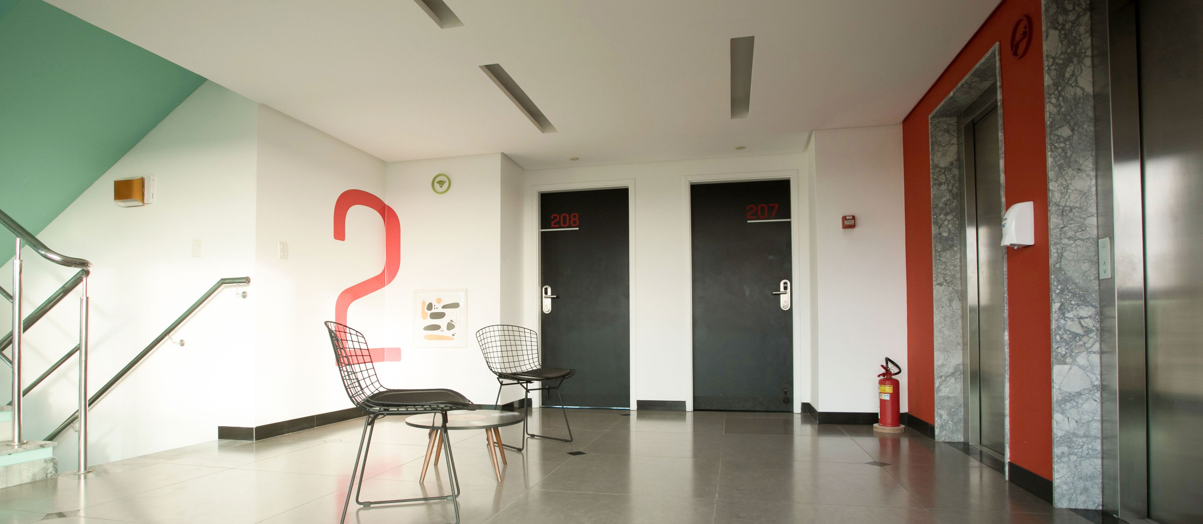 Um lobby moderno com uma escada, duas portas identificadas com os números 208 e 207, e um extintor de incêndio.