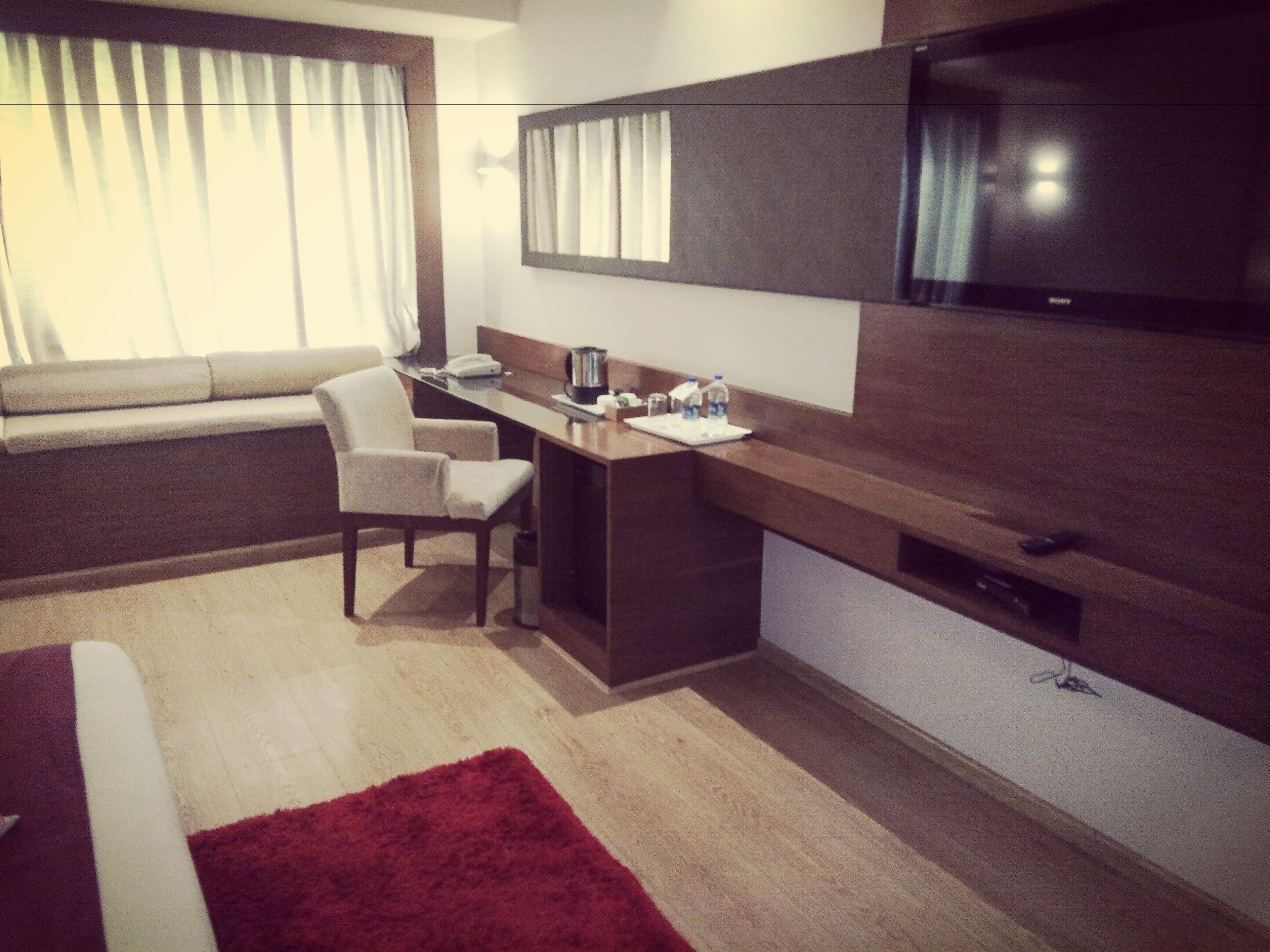 Quarto de hotel moderno com sofá, escrivaninha, cadeira, televisão de tela plana e uma grande janela com cortinas.