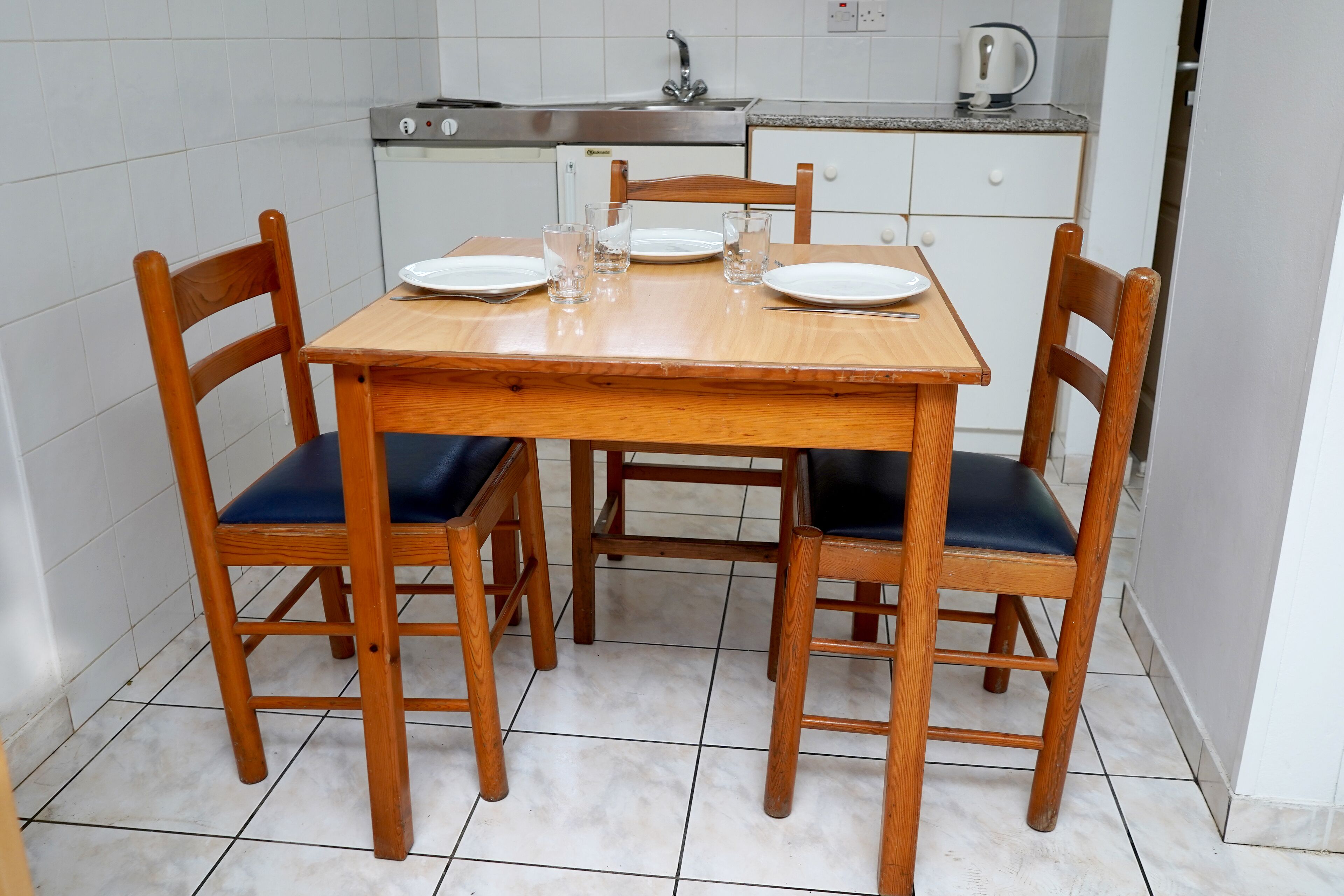 Uma mesa de jantar de madeira com quatro cadeiras, cada uma com uma almofada escura. A mesa está posta com pratos brancos, copos e uma faca.