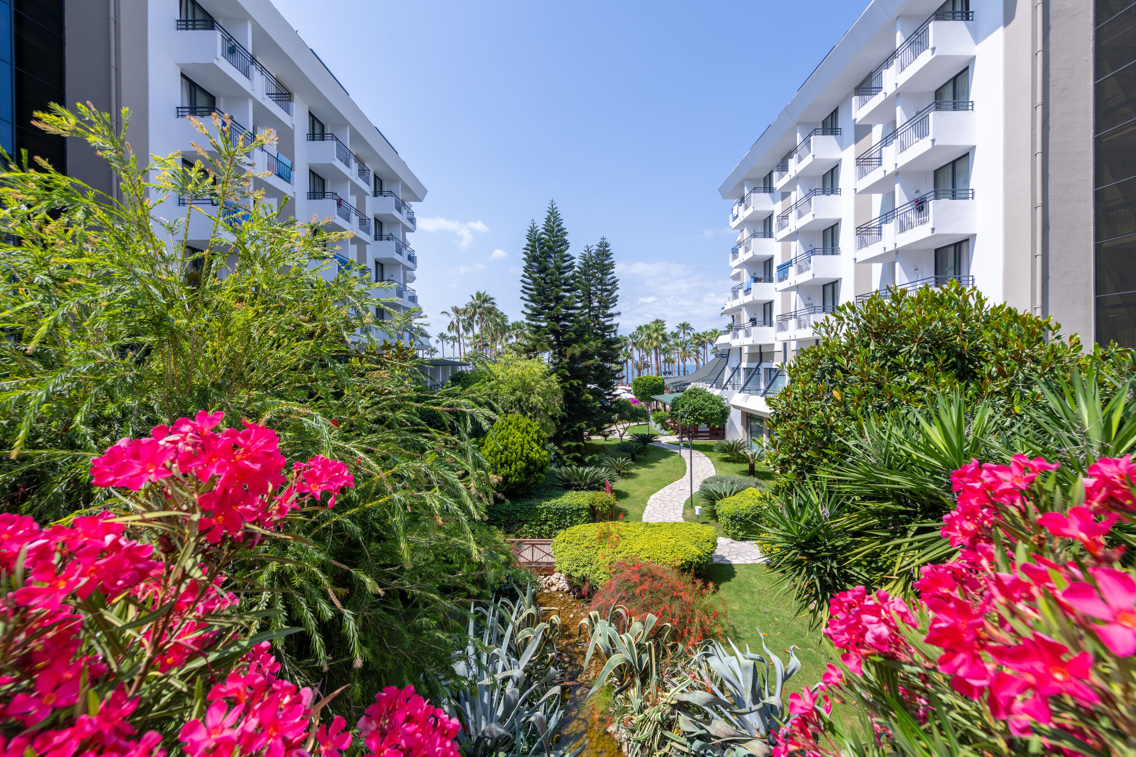 Um hotel moderno com um pátio central, apresentando um caminho, vegetação exuberante e plantas floridas.