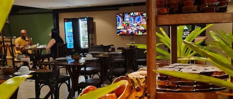 Um restaurante com mesas, cadeiras, uma tela de televisão e diversos pratos, incluindo pizza e uma bebida.