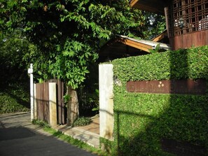 Uma entrada protegida por um muro de madeira e um portão de metal, rodeada por vegetação exuberante e uma sebe bem cuidada.