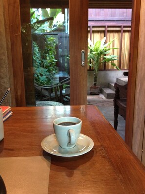Mesa de madeira com uma chávena de café num pires, porta que dá para um jardim com plantas e uma vedação de madeira.