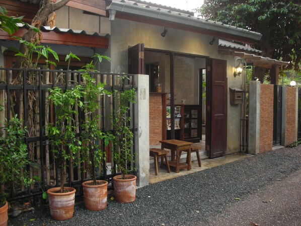 Um pequeno edifício rústico com porta de madeira e parede de tijolos. Há duas plantas em vasos na frente da porta.