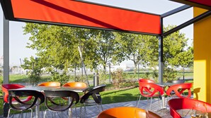 Uma área de refeições ao ar livre com cadeiras vermelhas e pretas, uma mesa e um ambiente envidraçado com vista para um gramado verde e árvores.