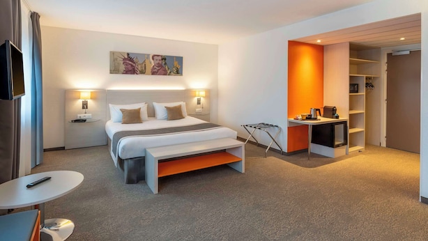 Quarto de hotel moderno com uma cama grande, mesas de cabeceira com luminárias, uma pequena escrivaninha com cadeira e um guarda-roupa com prateleiras.