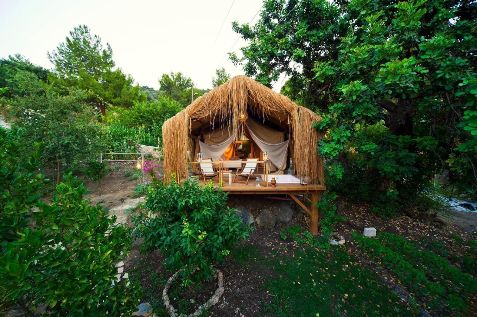Uma tenda rústica com telhado de palha e deck de madeira, rodeada por vegetação exuberante.