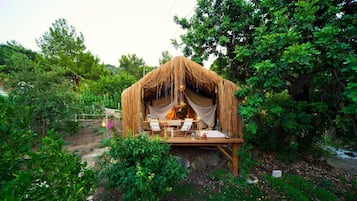 Uma tenda rústica com telhado de palha e deck de madeira, rodeada por vegetação exuberante.