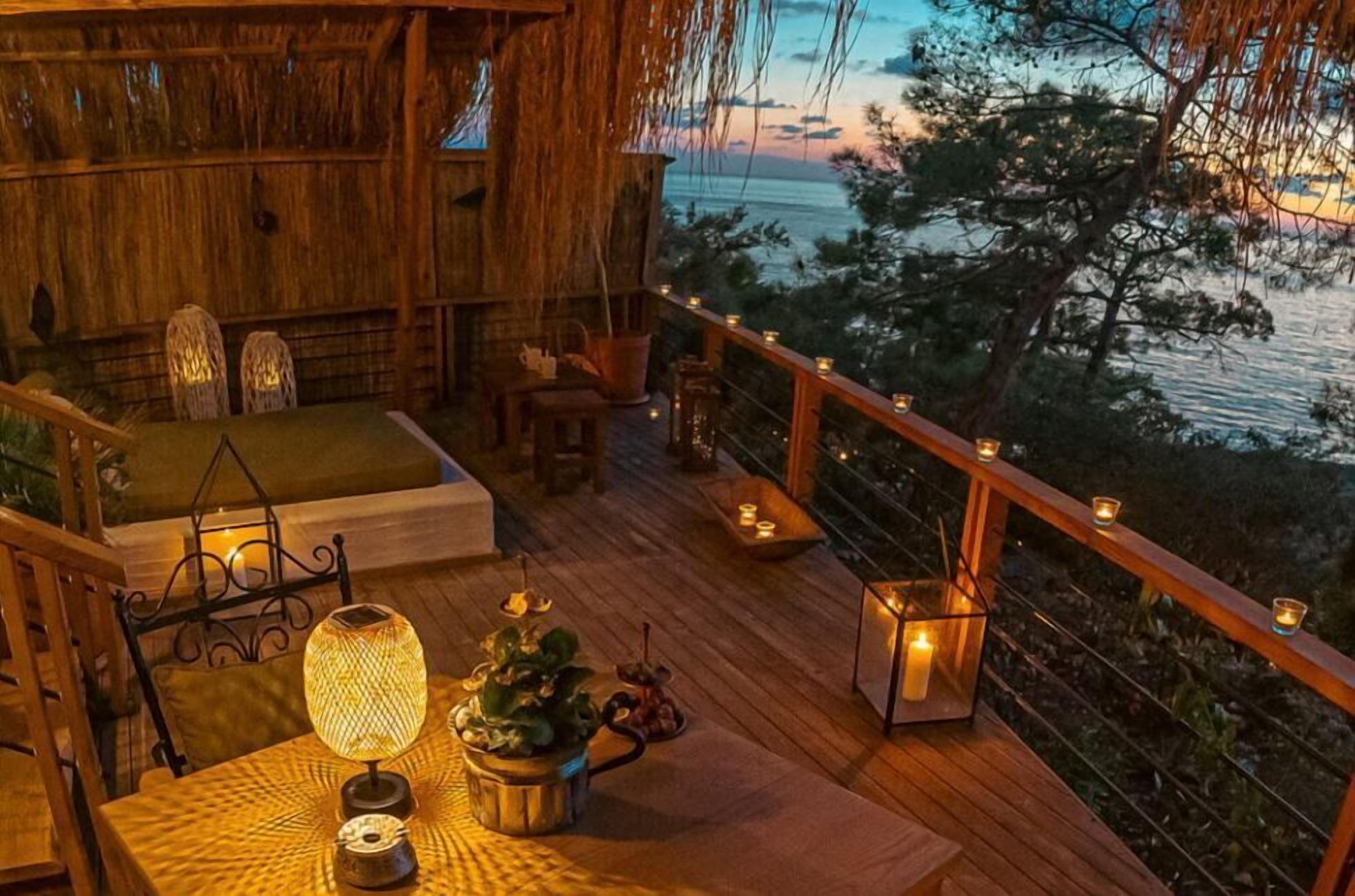 Um deck de madeira com vista para o oceano, área de estar com almofadas e uma mesa com candeeiro e velas.