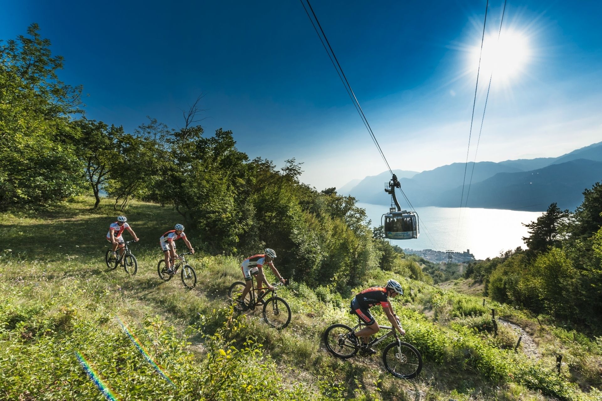 Um grupo de ciclistas numa trilha de montanha, com um teleférico ao fundo.