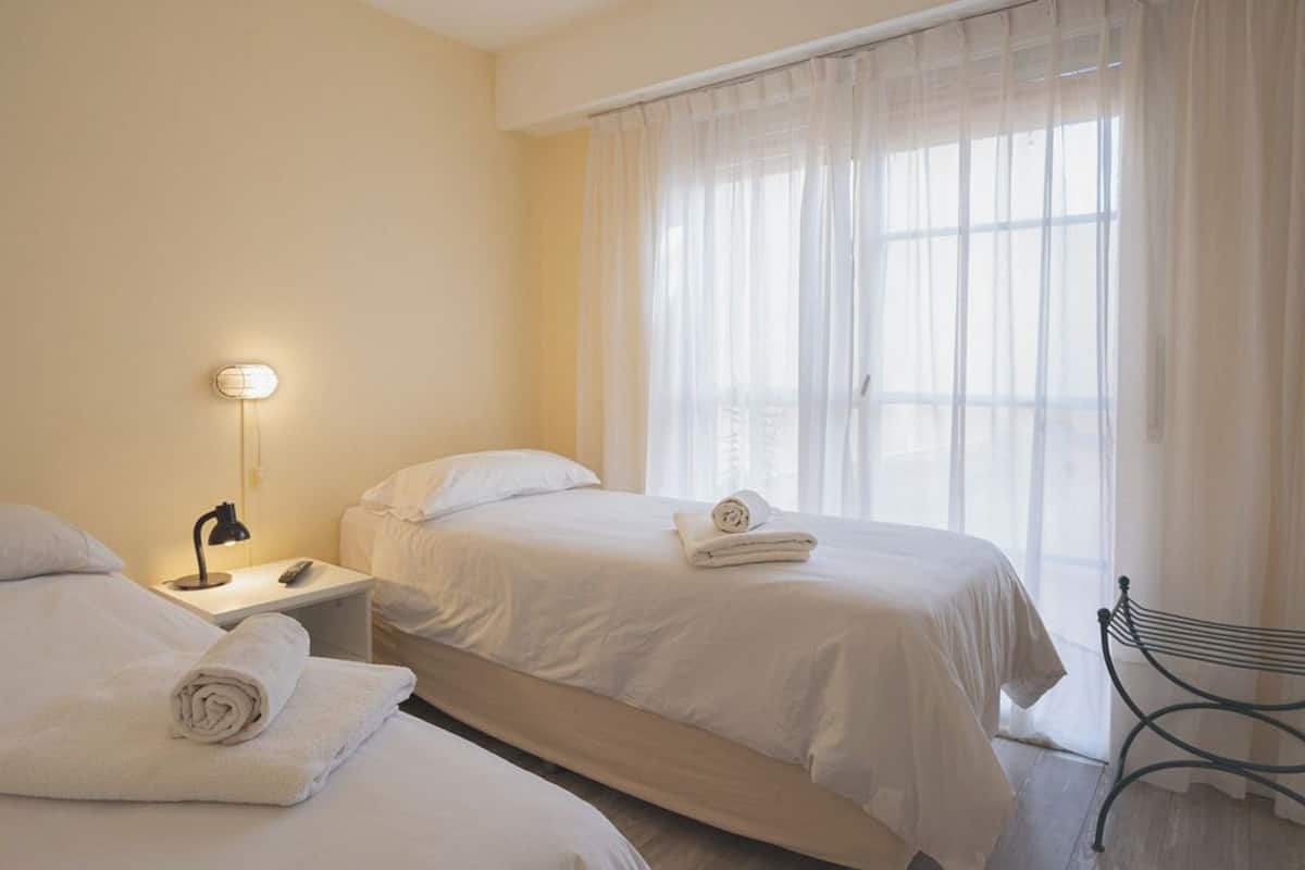 Quarto de hotel com duas camas, mesa de cabeceira com abajur, uma cadeira e uma janela grande com cortinas leves.