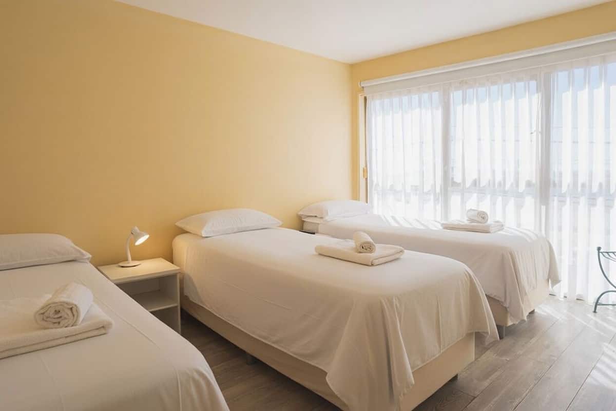Quarto de hotel com duas camas, mesa de cabeceira com abajur e uma janela grande com cortinas leves.