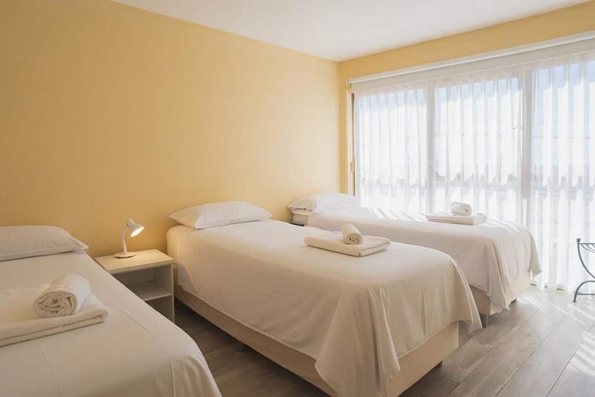 Quarto de hotel com duas camas, uma mesa de cabeceira com abajur e uma janela com cortinas translúcidas.