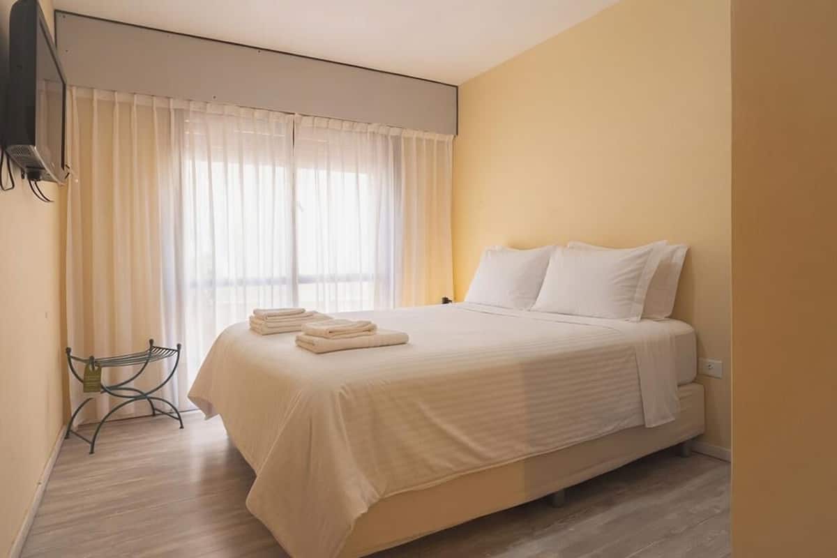 Quarto de hotel com cama, janela com cortinas, mesa de cabeceira e televisão fixada na parede.