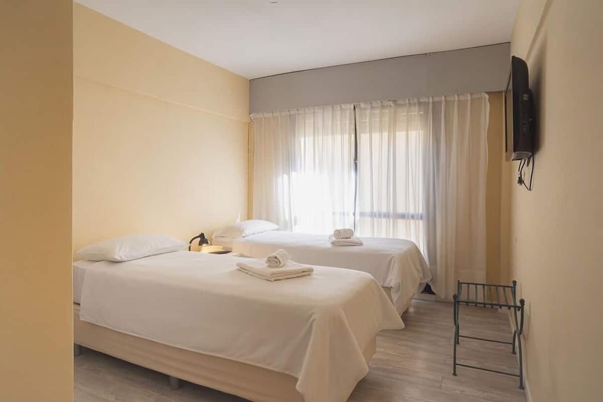 Quarto de hotel com duas camas, uma televisão presa na parede e uma janela com cortinas leves.