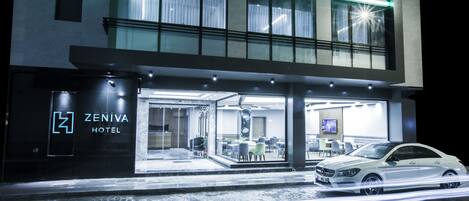 Hotel moderno com fachada de vidro, entrada visível e um carro estacionado na frente.