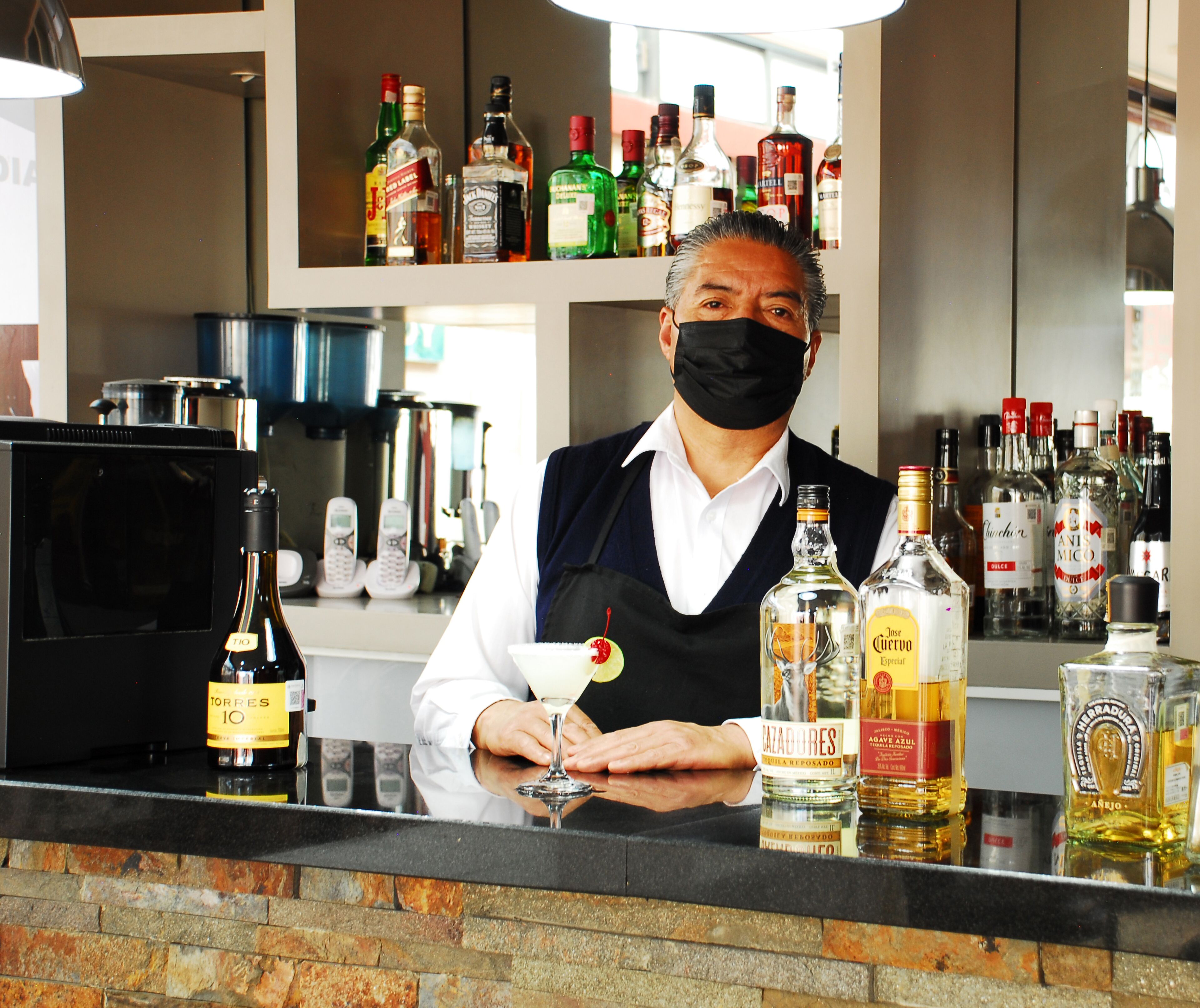 Uma área de bar com um bartender, diversas garrafas de bebidas alcoólicas e uma máquina de café.