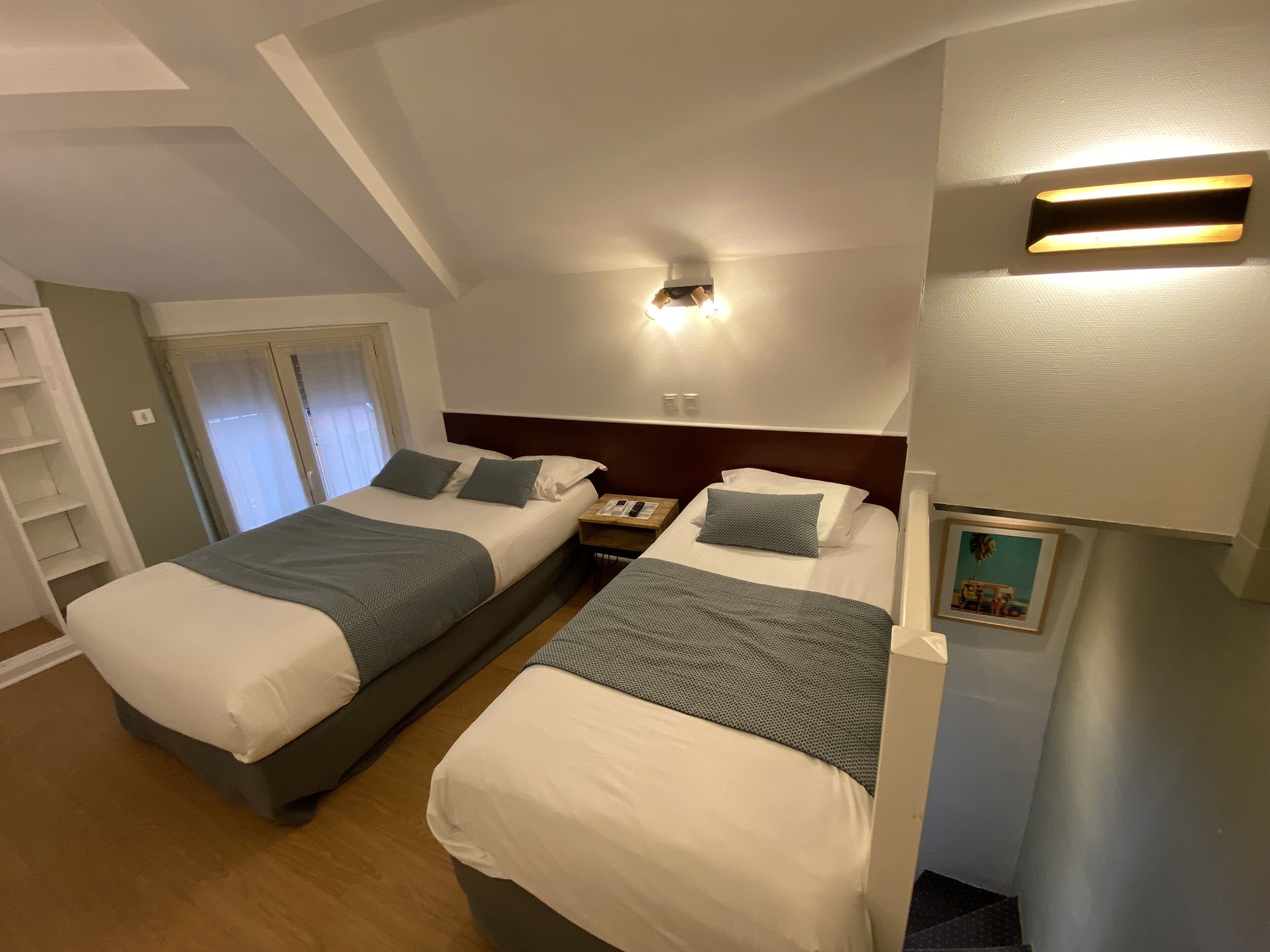 Quarto de hotel com duas camas, uma mesinha de cabeceira, uma luminária na parede e um quadro emoldurado.
